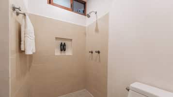 Pequeno espaço de duche fechado, com azulejos bege, sanita branca e suporte para toalhas.