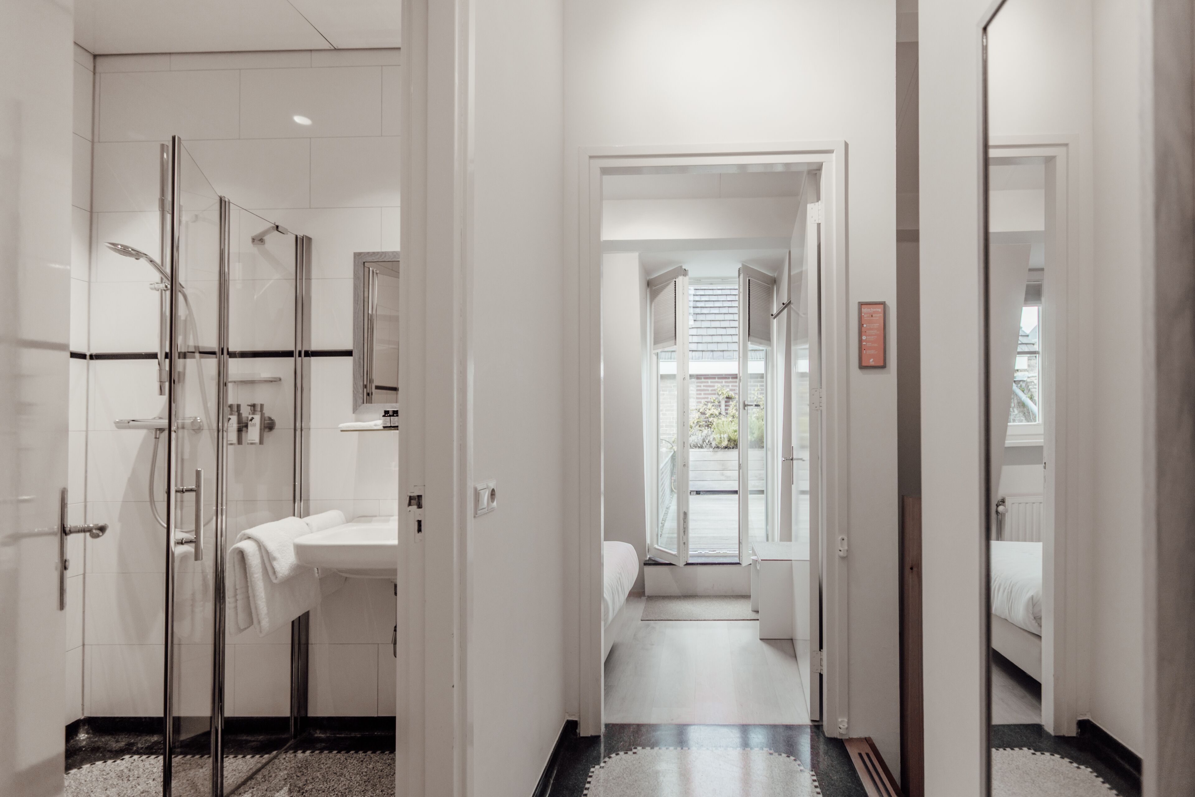 Quarto de hotel moderno com chuveiro de vidro, pia branca e cama com vista para o exterior.