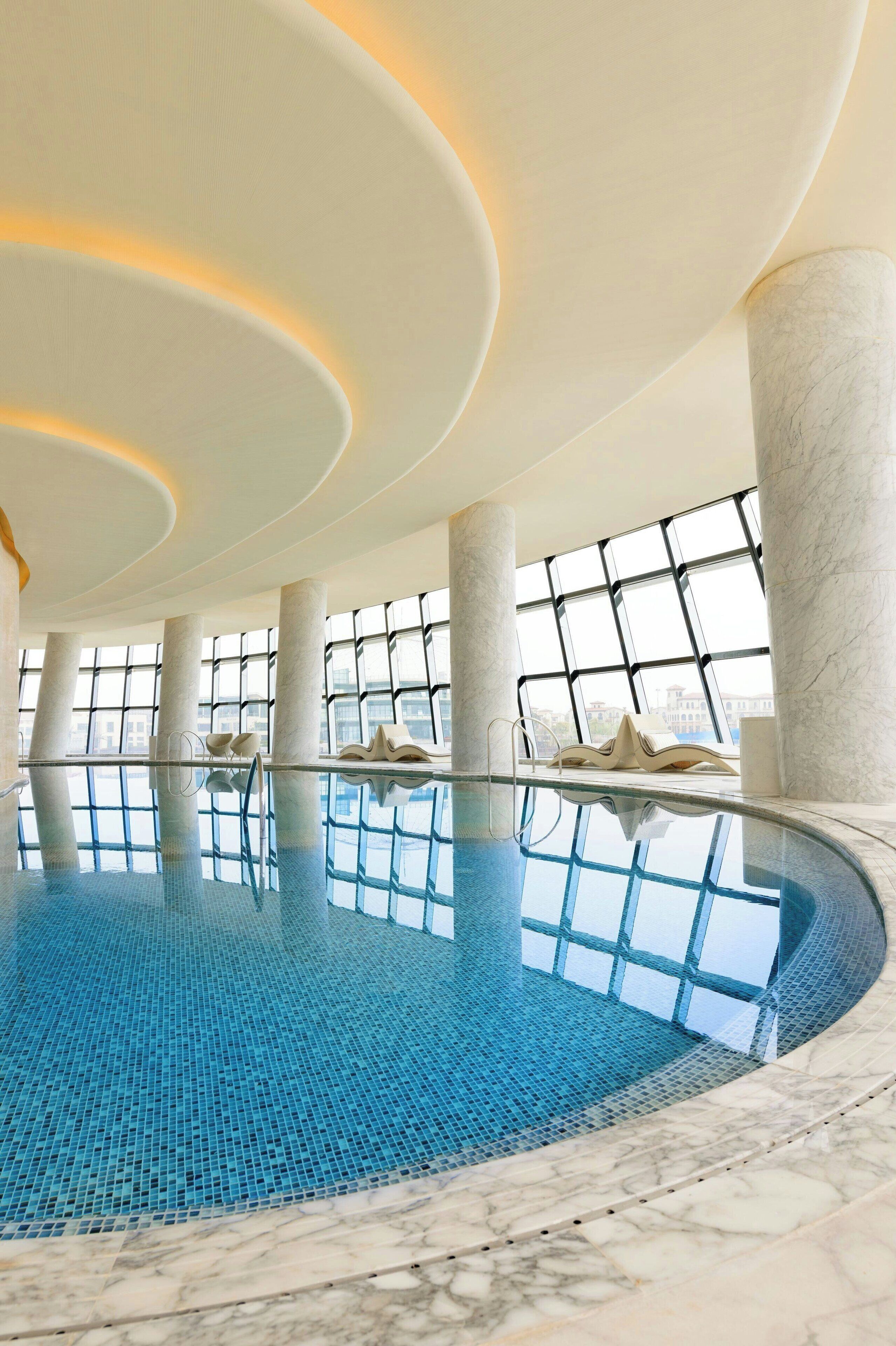 Uma piscina interior com água azul cristalina, rodeada por colunas de mármore e amplas janelas.