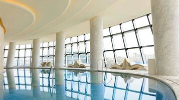 Uma piscina interior com água azul cristalina, rodeada por colunas de mármore e amplas janelas.