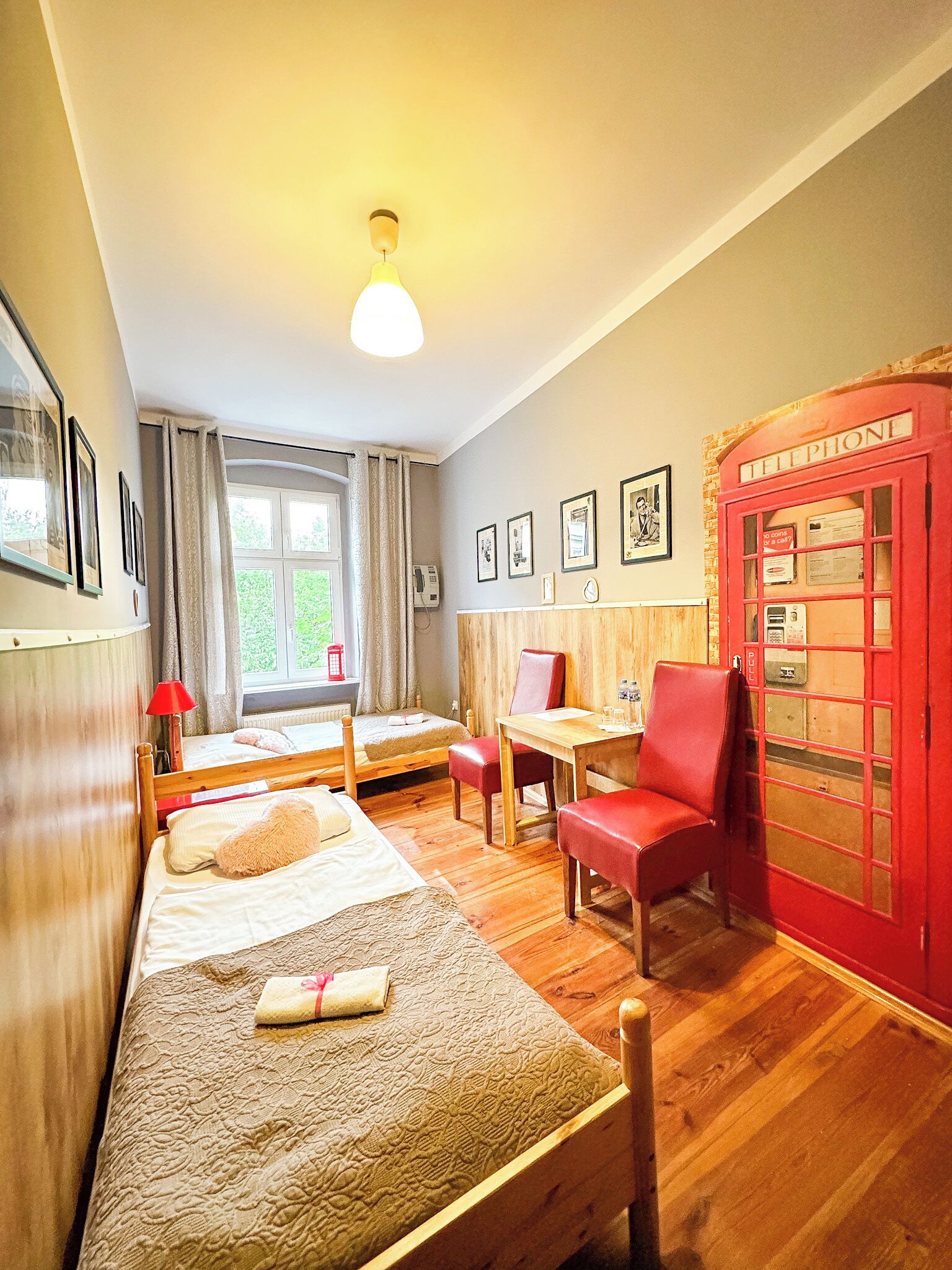 Quarto com duas camas de madeira, um telefone público vermelho, uma mesa de madeira e uma janela com cortinas.