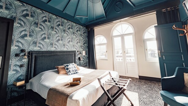 Quarto com cama grande, cabeceira, escrivaninha e cadeira. Possui duas janelas e uma porta.
