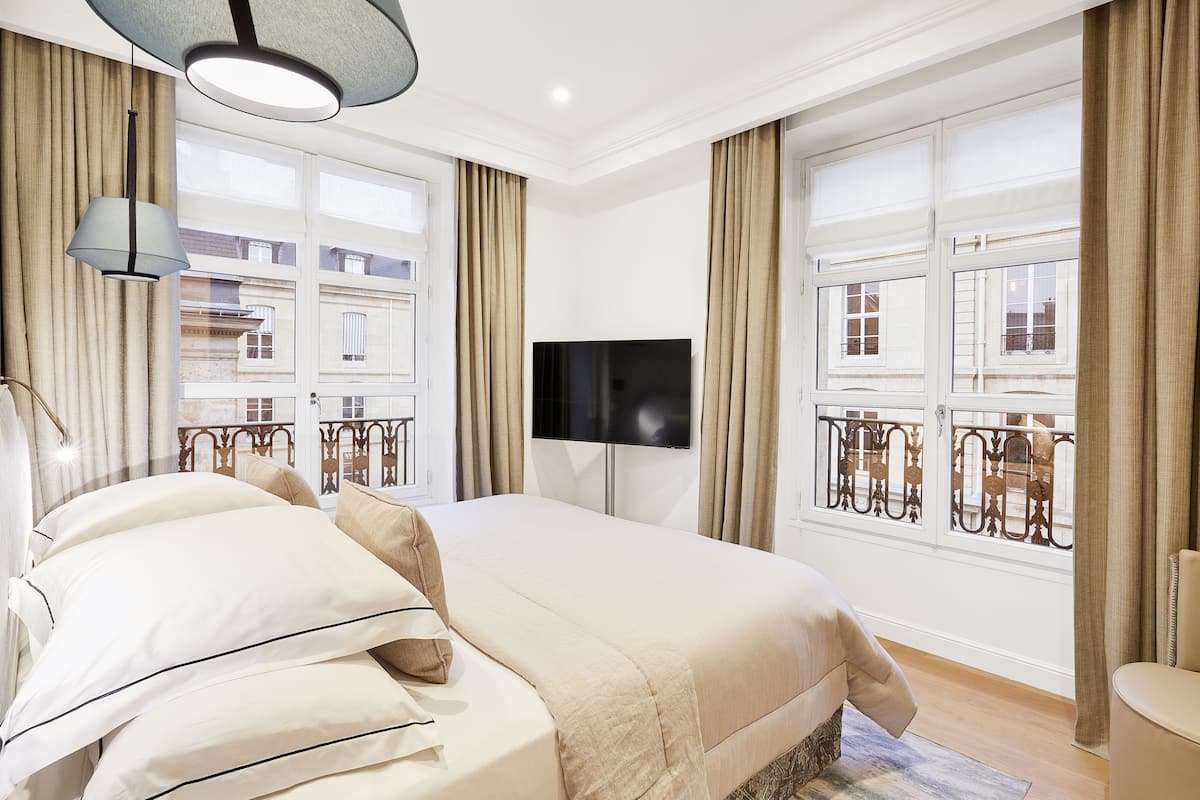 Quarto de hotel com uma cama, televisão e janelas amplas com cortinas.