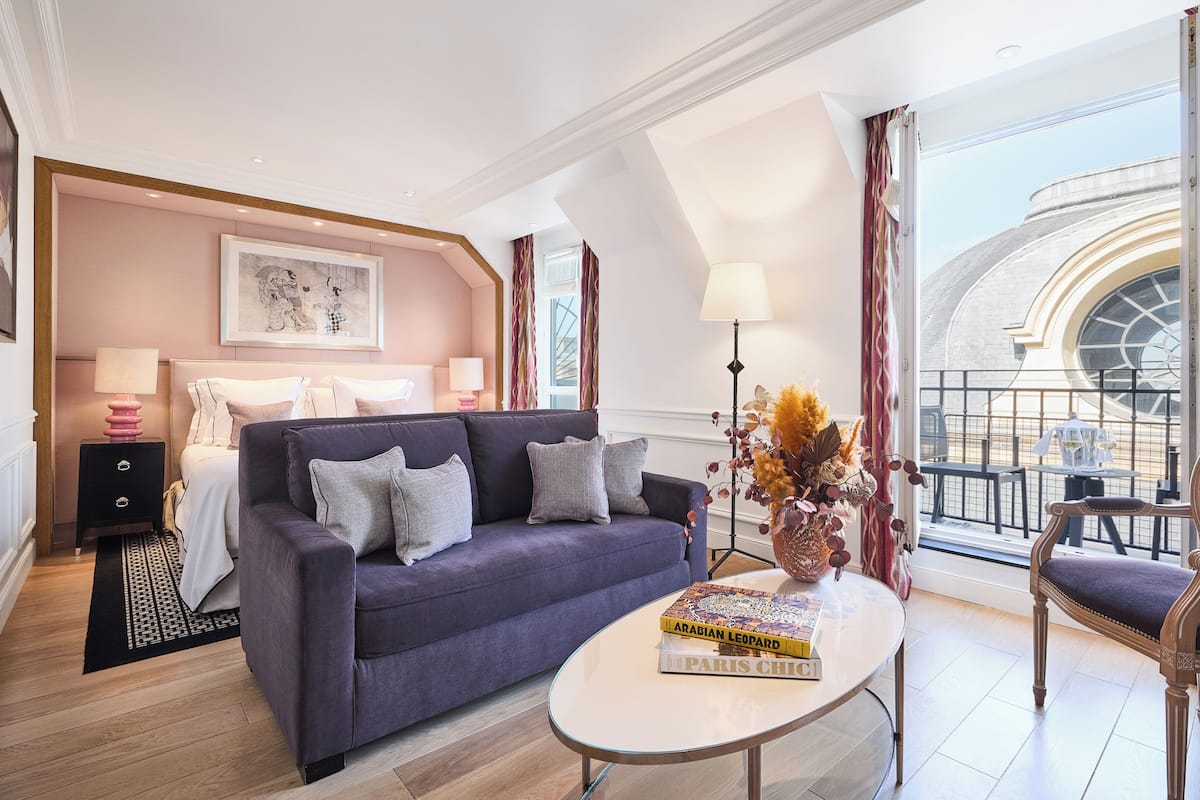 Quarto de hotel moderno com sofá, uma mesinha com livros, cama com mesa de cabeceira e varanda com vista.
