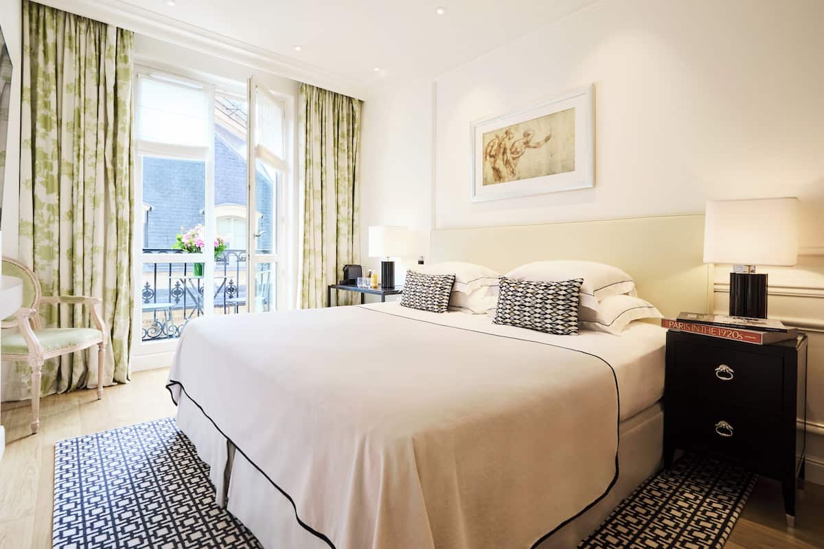Quarto de hotel com uma cama grande, uma mesinha de cabeceira com abajur, uma pequena mesa com cafeteira, uma cadeira e vista para o exterior.