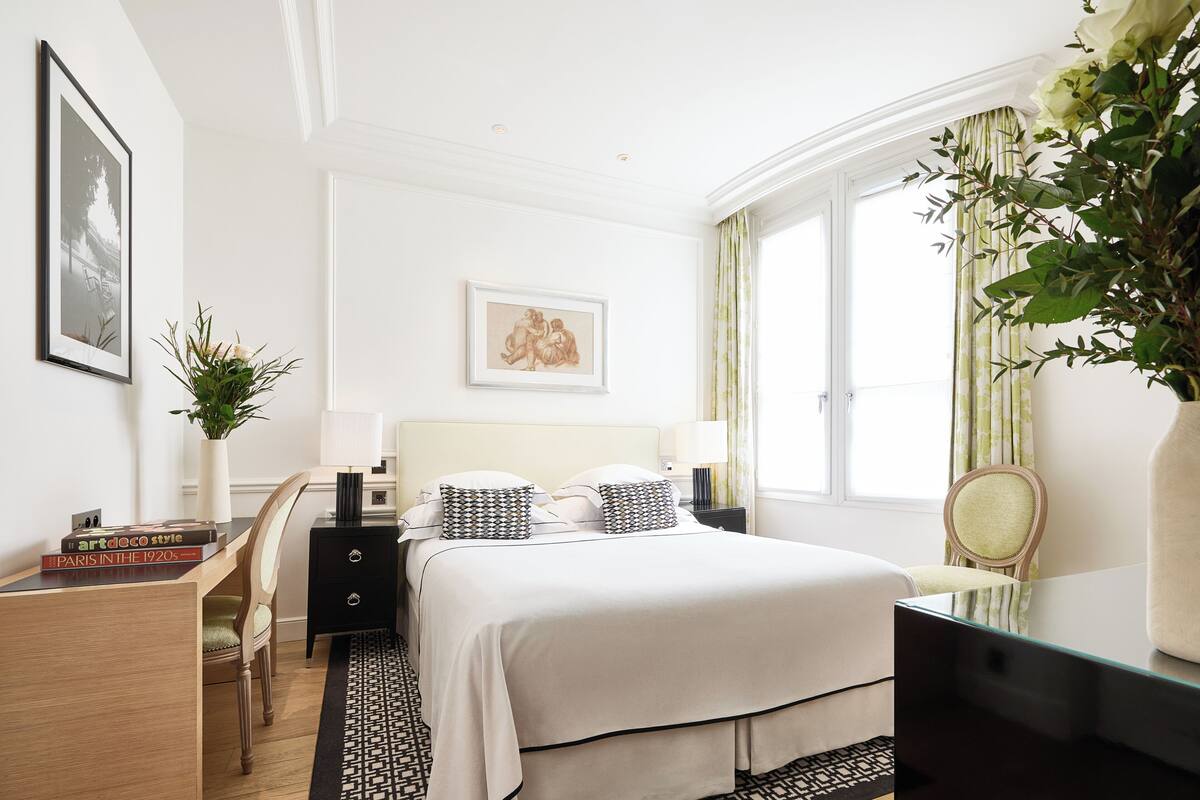 Quarto de hotel com cama, mesas de cabeceira, escrivaninha, cadeira, janela com cortinas e um quadro na parede.