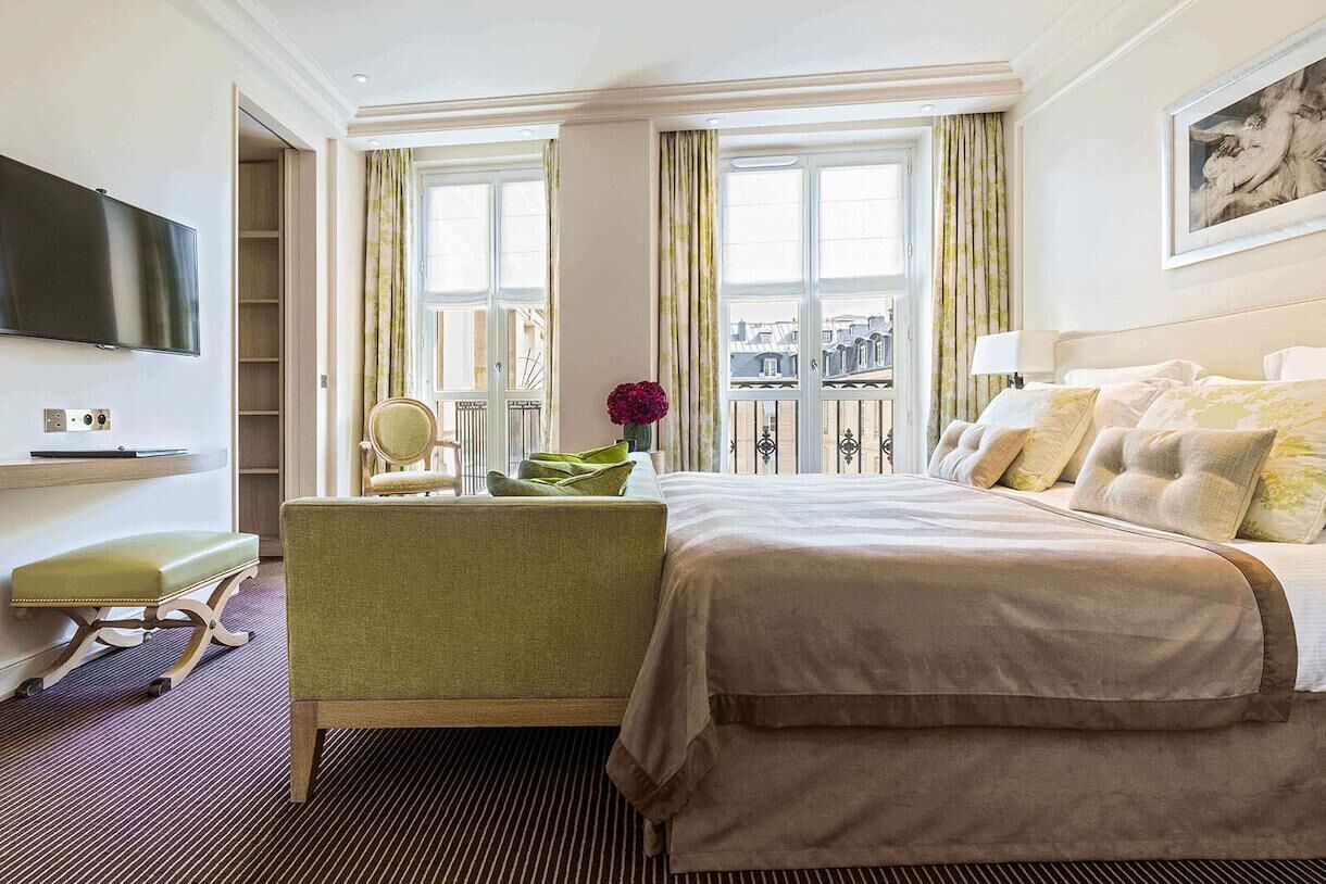 Quarto de hotel com cama, uma cadeira, televisão, janela com cortinas e vista para a varanda.