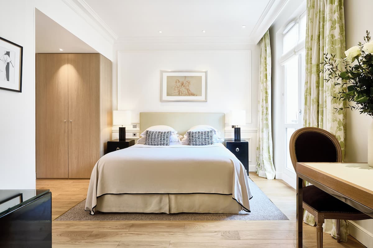 Quarto de hotel com cama, mesas de cabeceira, escrivaninha, cadeira, guarda-roupa e janela com cortinas.