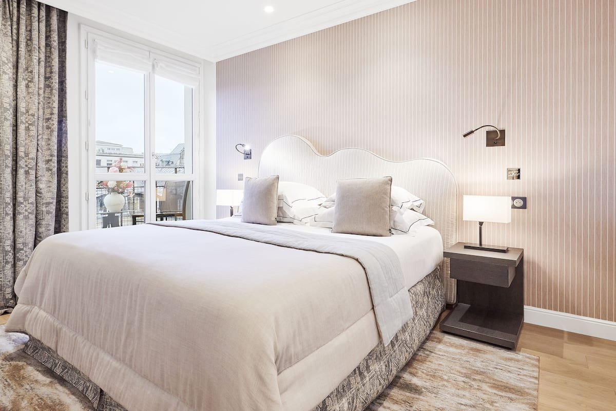 Quarto moderno com uma cama grande, mesa de cabeceira com abajur e vista para a cidade pela janela.