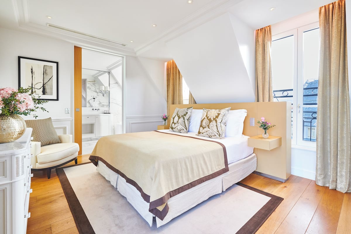 Quarto com cama grande, mesa de cabeceira, criadeira, cadeira, cômoda e janela com cortinas.