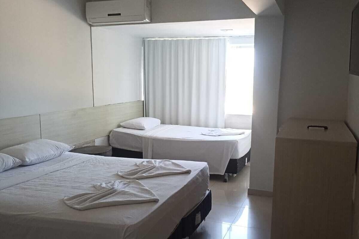 Quarto de hotel com duas camas, uma mesinha, um armário e uma janela com cortinas.
