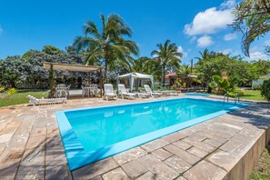 Uma piscina retangular com deck de pedra, rodeada por espreguiçadeiras brancas e uma área de estar coberta.