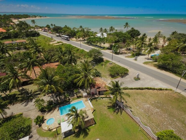 Vista aérea de uma propriedade costeira com piscina, estrada e praia ao fundo.