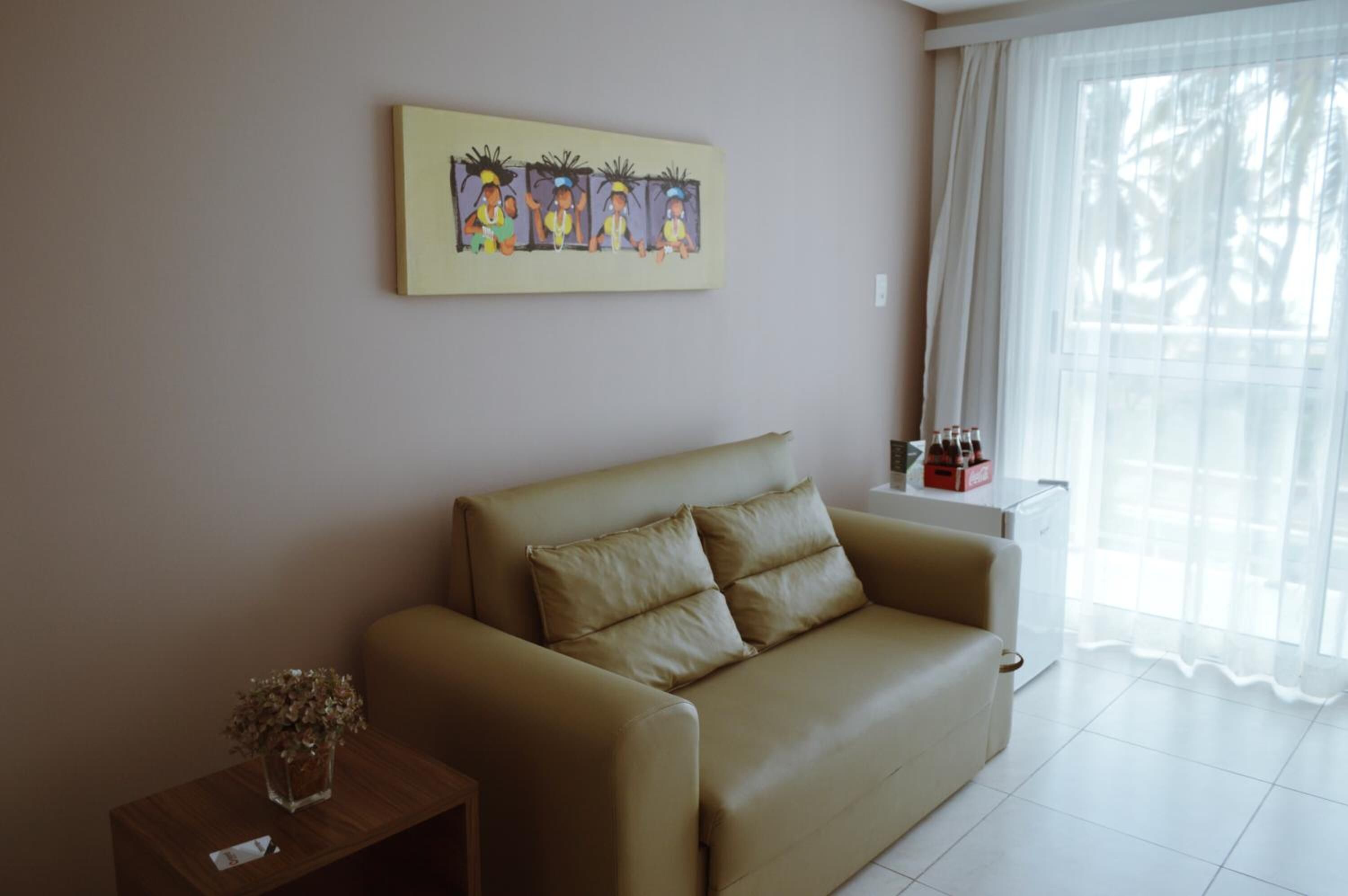 Uma sala de estar com um sofá bege, uma mesinha com um vaso, um quadro na parede e uma janela com cortinas leves.