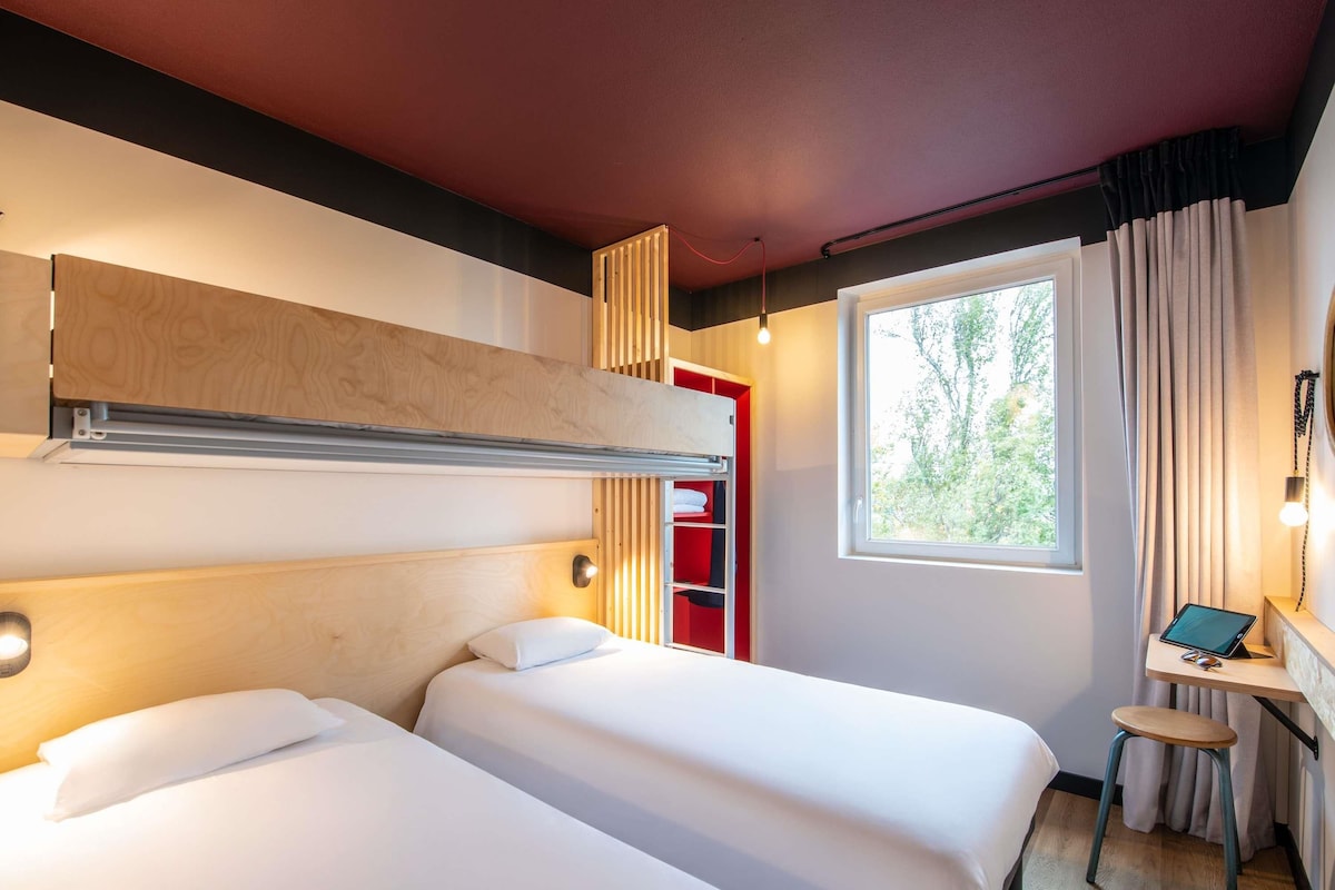 Quarto de hotel com duas camas, uma mesa com cadeira, uma janela com cortinas e um móvel de armazenamento vermelho.