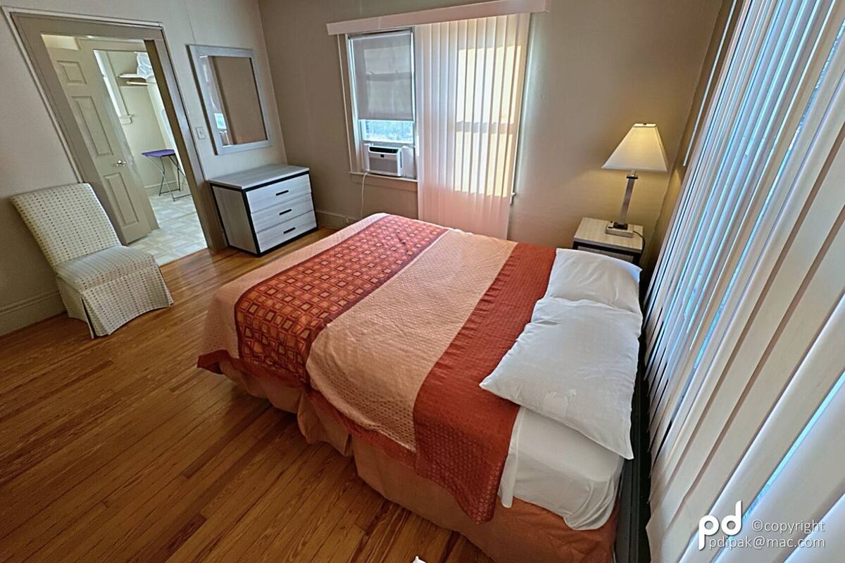Um quarto com cama, mesa de cabeceira, luminária, cadeira, cômoda, janela com persianas e porta.