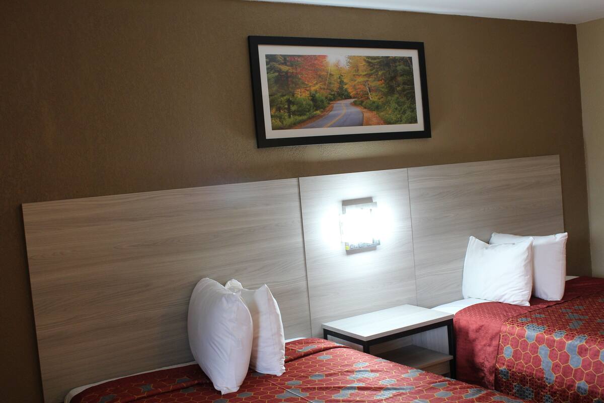 Quarto de hotel com cabeceira de madeira, cama com roupa de cama vermelha, duas almofadas brancas, mesa de cabeceira e um quadro com uma estrada por uma floresta.