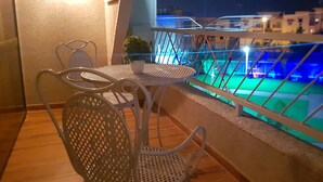 Varanda com mesa e cadeiras, um vaso de planta e vista para a área da piscina iluminada à noite.