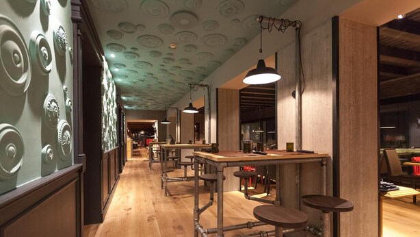 Uma área de bar com piso de madeira, banquetas de metal e iluminação pendente.