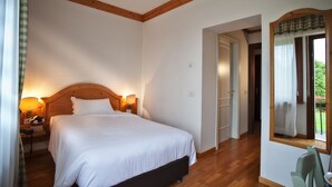 Quarto com cabeceira de madeira, cama com roupa de cama branca, luminárias de cabeceira, um espelho e vista para um jardim pela janela.
