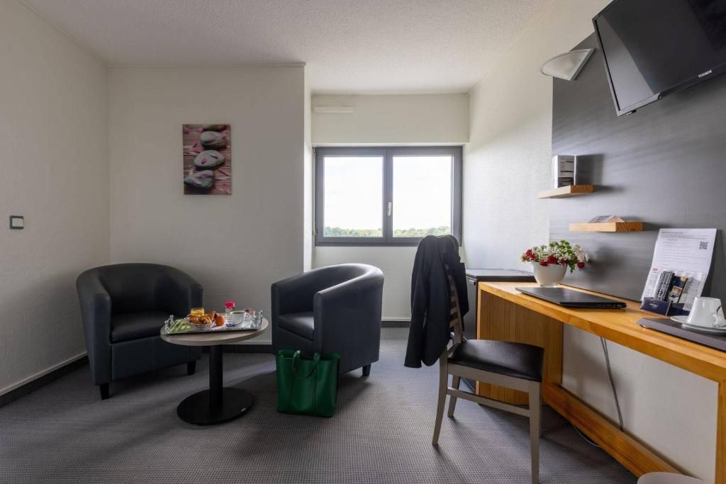 Quarto de hotel com duas poltronas, uma mesa pequena com bandeja de comida, escrivaninha com computador, TV fixada na parede e janela com vista para o verde.