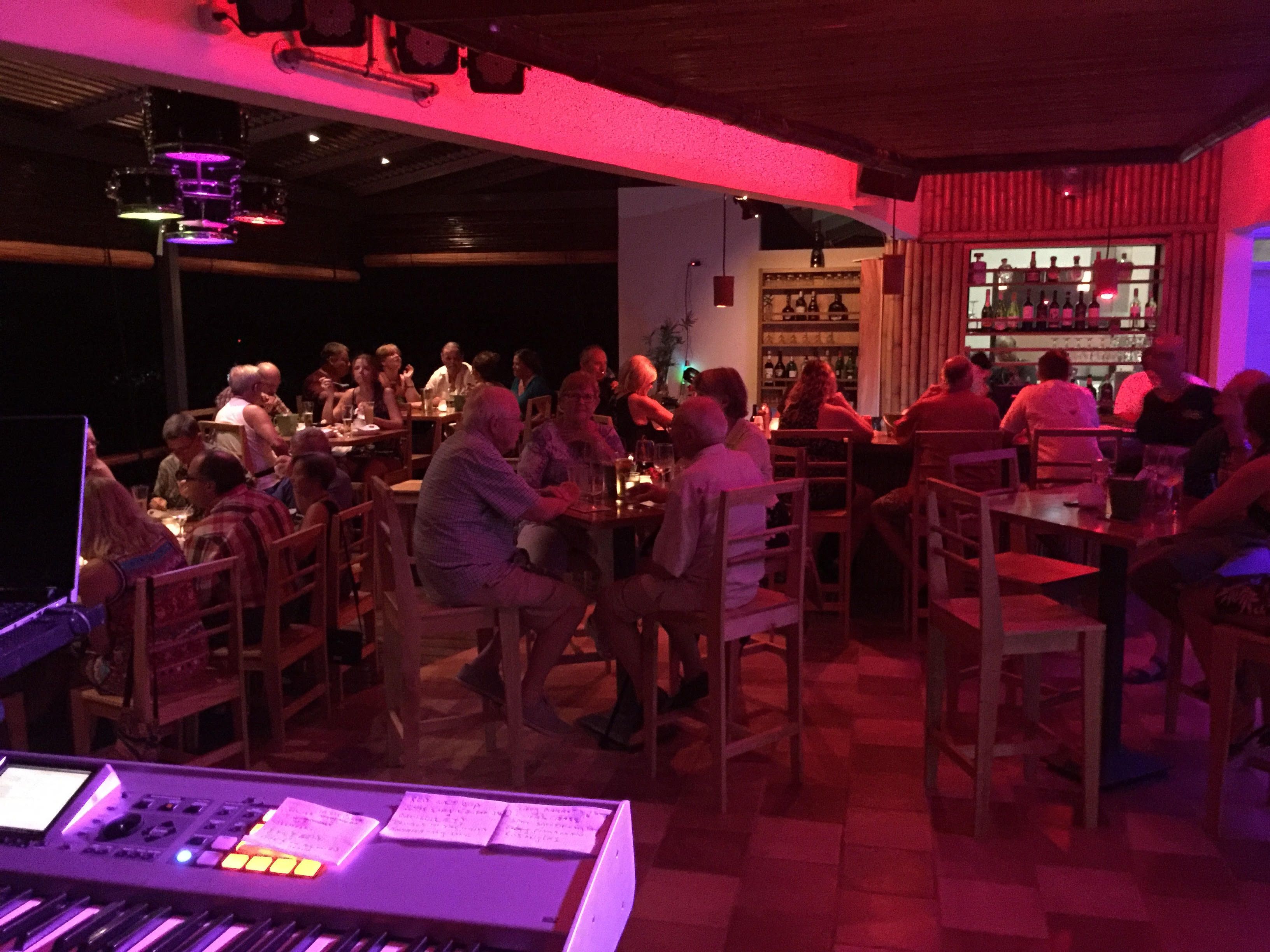 Restaurante com clientes sentados em mesas, área de bar com garrafas e equipamento de DJ em primeiro plano.