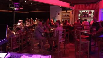 Restaurante com clientes sentados em mesas, área de bar com garrafas e equipamento de DJ em primeiro plano.