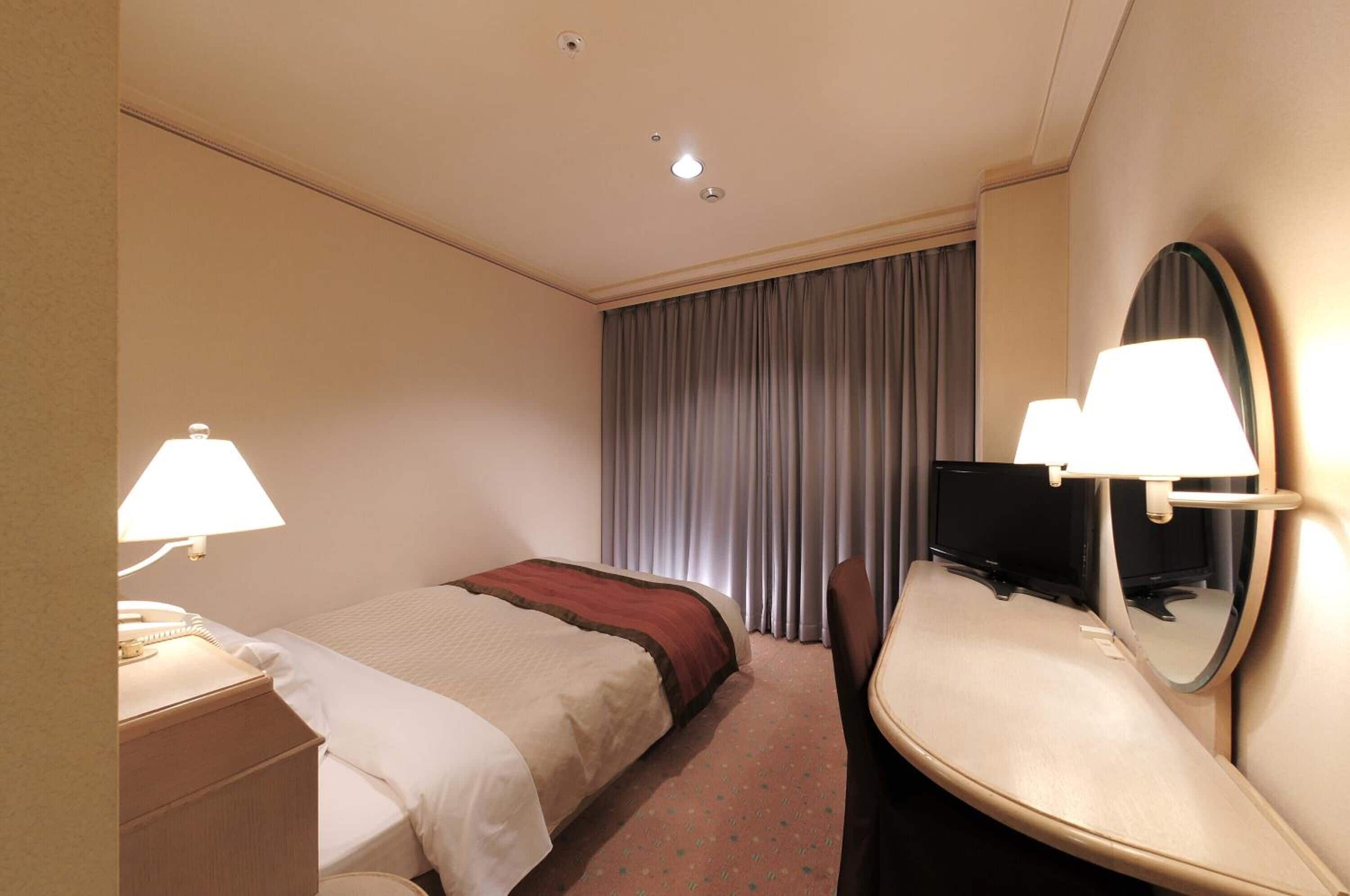 Quarto de hotel com cama, mesa de cabeceira, luminária, espelho e cortinas.