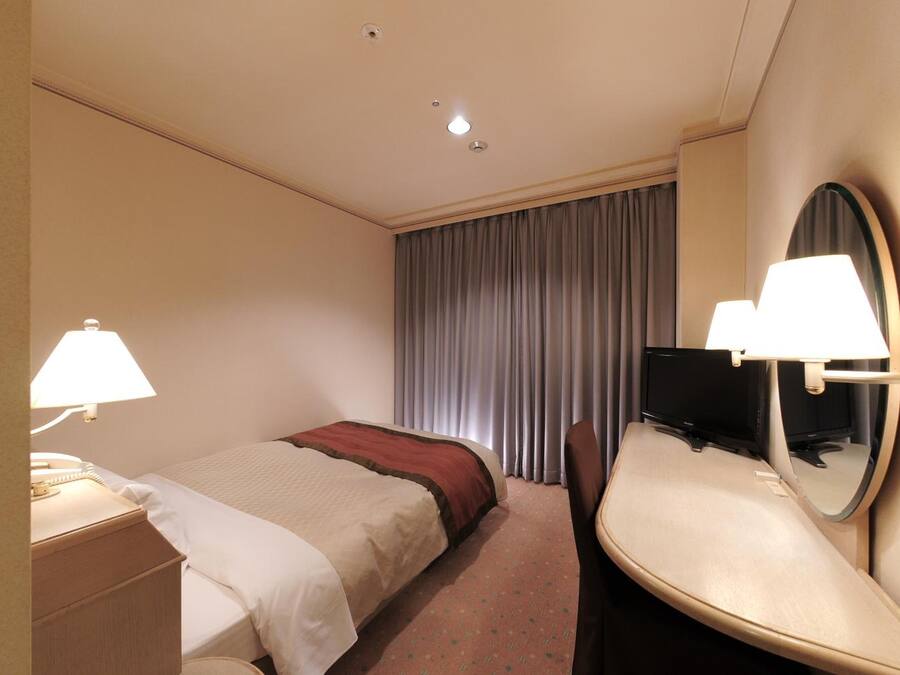 Quarto de hotel com cama, mesa de cabeceira, luminária, espelho e cortinas.