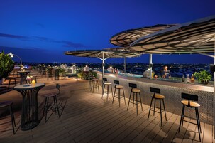 Uma área de bar ao ar livre com piso de madeira, banquetas e guarda-sóis para proteção do sol.