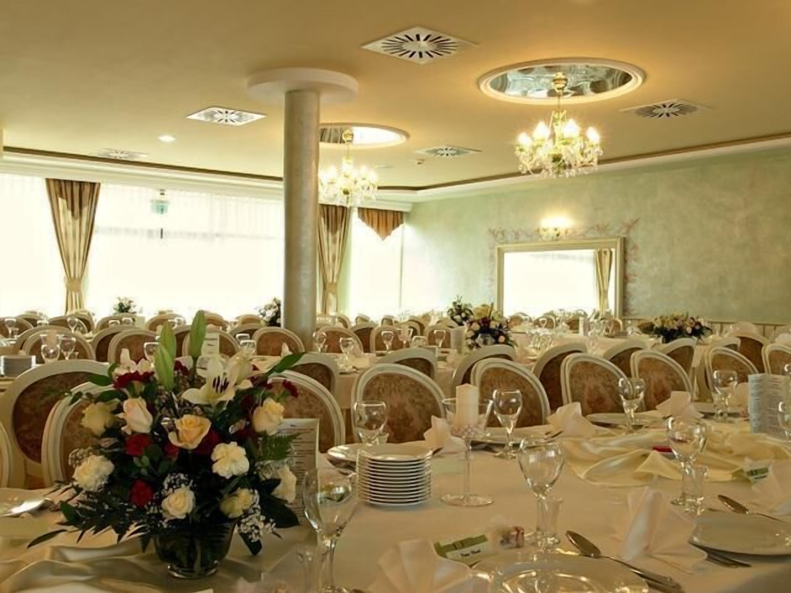 Uma sala de banquetes com mesas redondas dispostas para refeições, cada uma com toalhas brancas, pratos e taças de vinho.