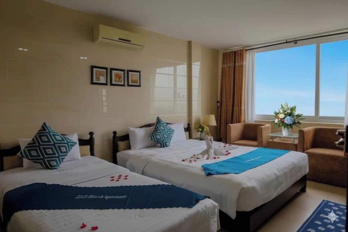 Quarto de hotel com duas camas, área de estar com um sofá e poltronas, uma mesinha de centro com um vaso, e uma janela grande com vista.