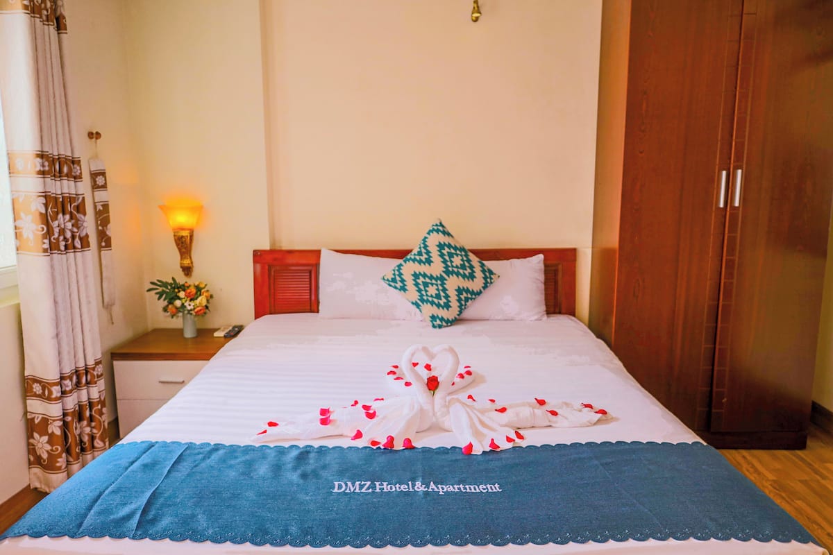 Quarto de hotel com uma cama bem arrumada, uma mesa de cabeceira com abajur e um guarda-roupa de madeira.