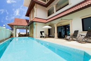 Piscina com espreguiçadeiras e guarda-sóis, área de varanda coberta e um edifício com telhado em azulejos e colunas.