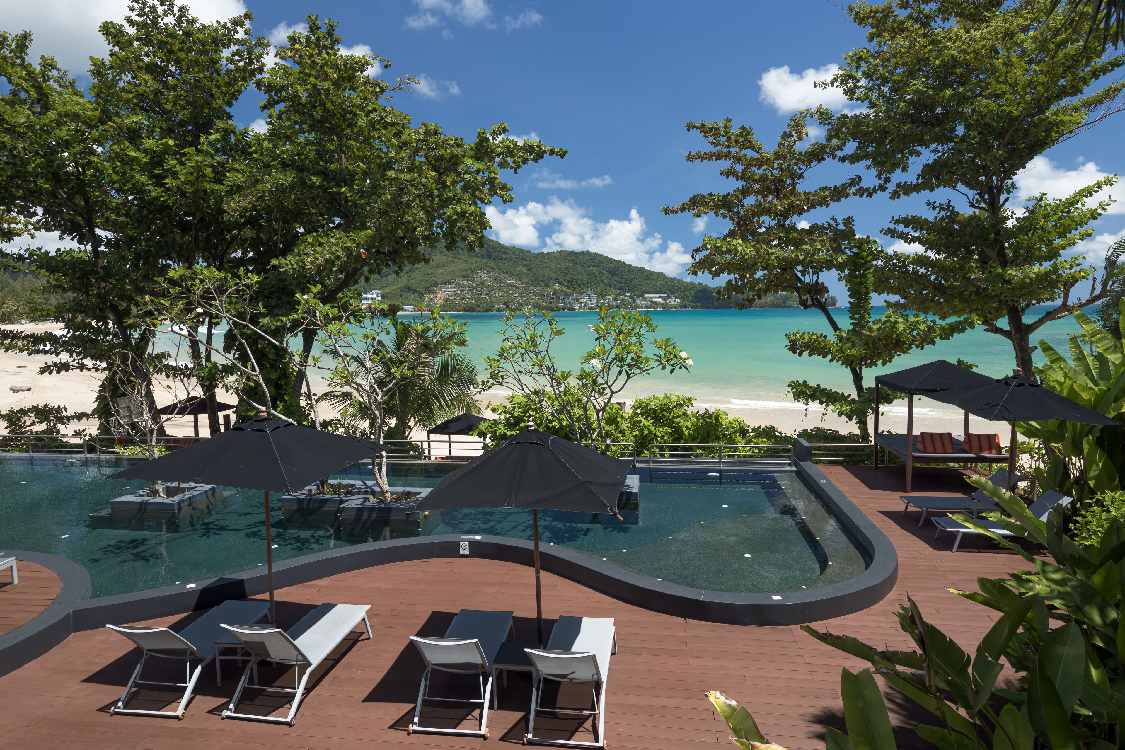 Área da piscina com espreguiçadeiras e guarda-sóis, com vista para uma praia e mar turquesa.