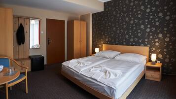 Quarto de hotel com uma cama de solteiro, escrivaninha com cadeira, guarda-roupa, mesa de cabeceira pequena com abajur e lixeira.