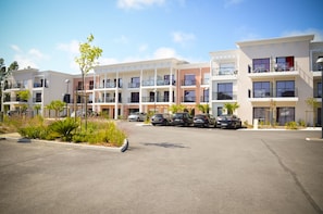 Um edifício residencial com vários andares, varandas, um estacionamento e uma área ajardinada com árvores e arbustos.