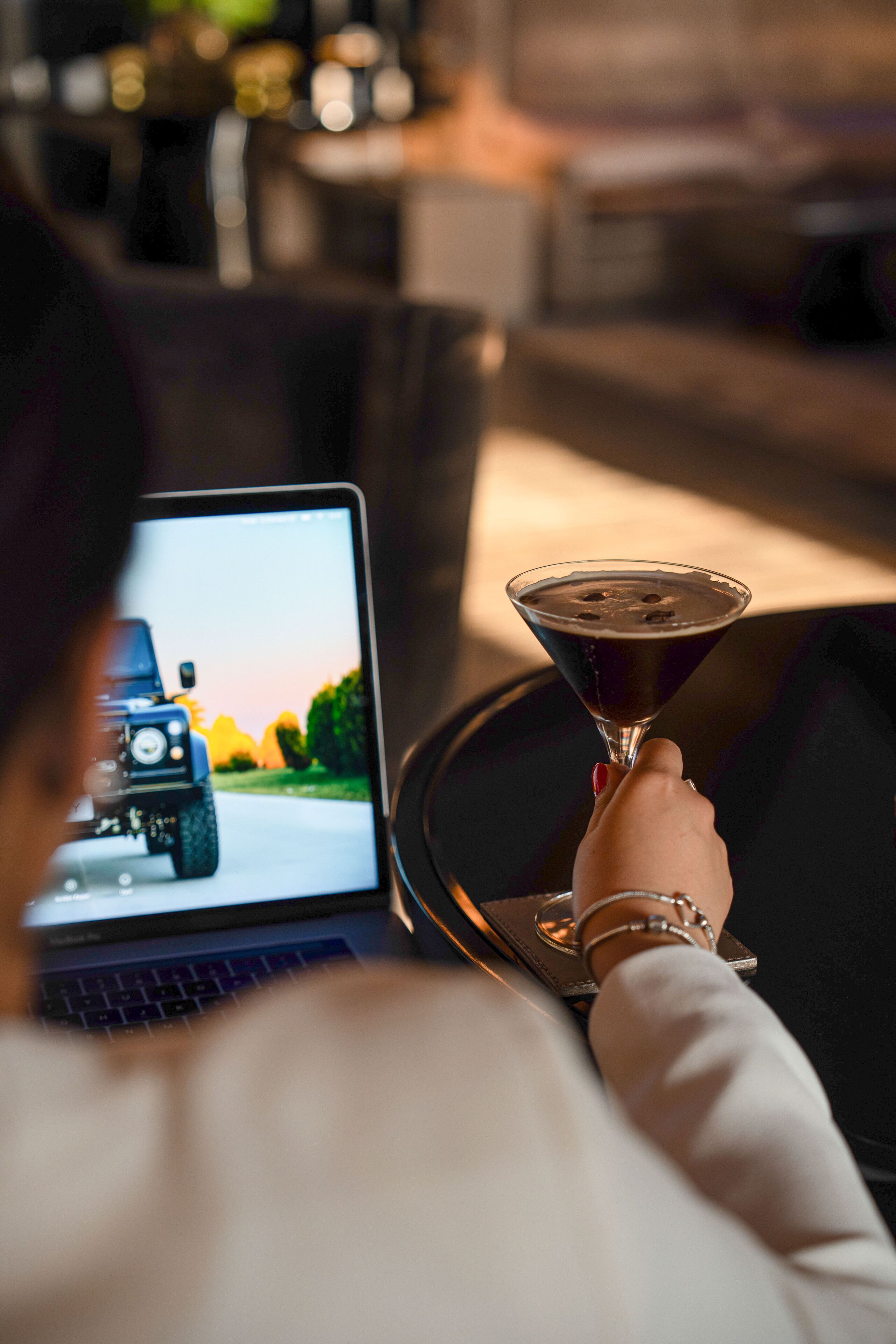 Uma pessoa segurando uma taça de martini com um laptop exibindo um carro na tela.