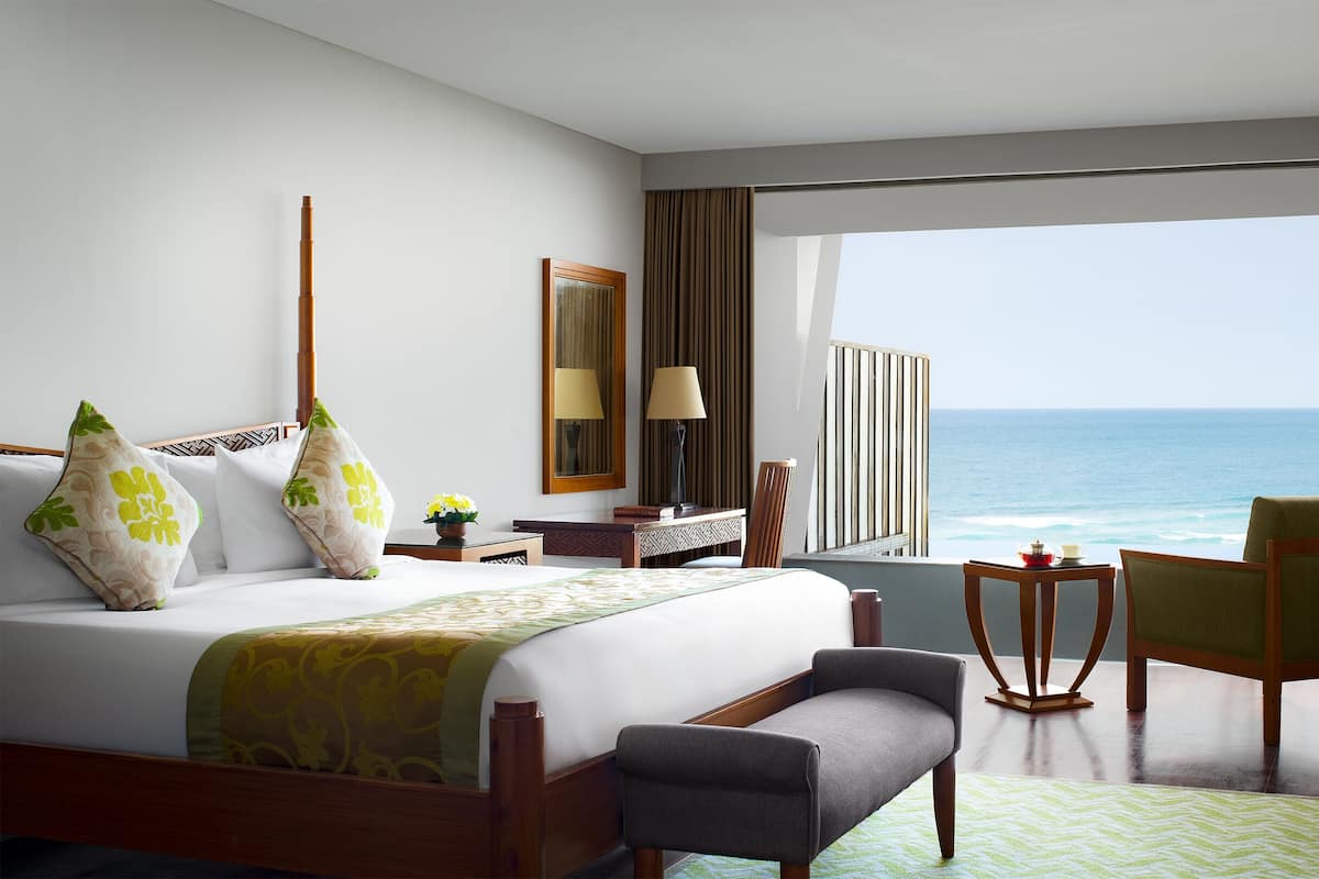 Quarto de hotel com uma cama grande, uma cadeira, uma mesa pequena, uma luminária e vista para o oceano.