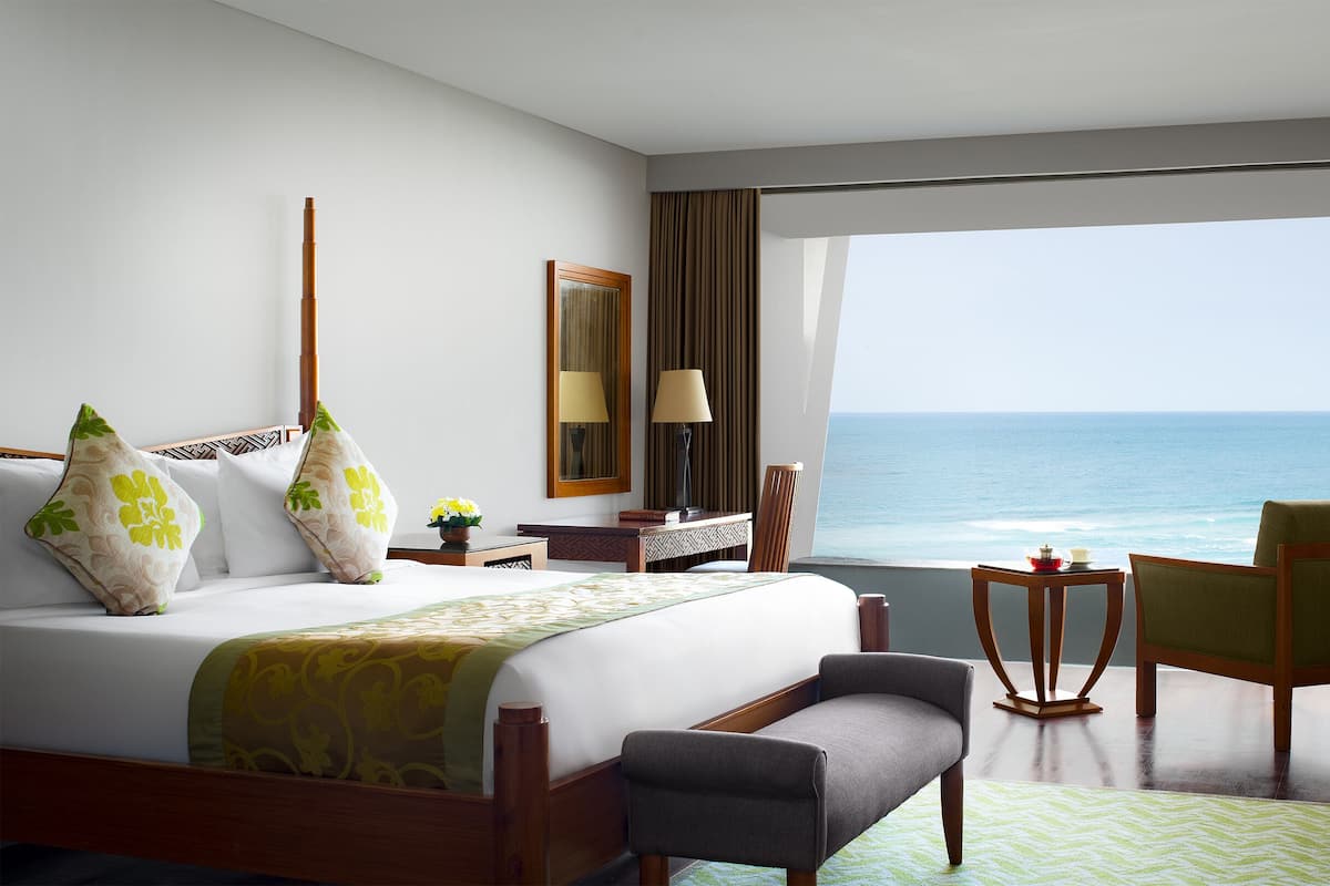 Quarto de hotel com uma cama grande, uma cadeira, uma mesinha, um espelho e vista para o oceano.