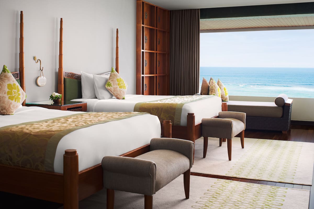 Quarto de hotel com duas camas, área de estar, vista para o oceano e uma janela grande.