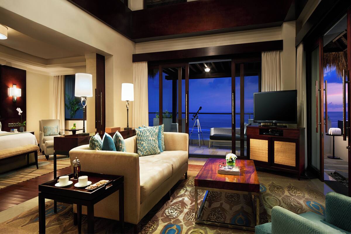 Quarto de hotel com sofá, poltronas, mesa de centro, televisão e varanda com vista para o oceano.