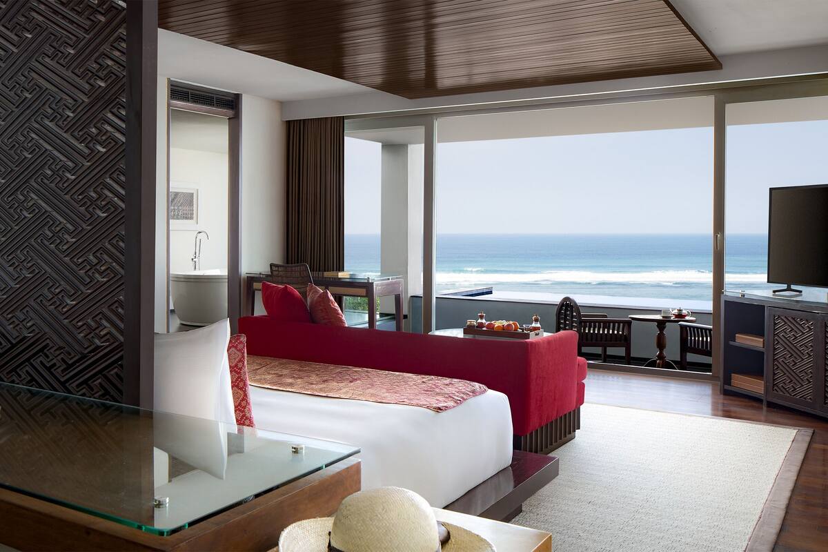 Quarto de hotel moderno com uma cama grande, uma mesa de vidro, vista para o mar e uma varanda com área de refeições.
