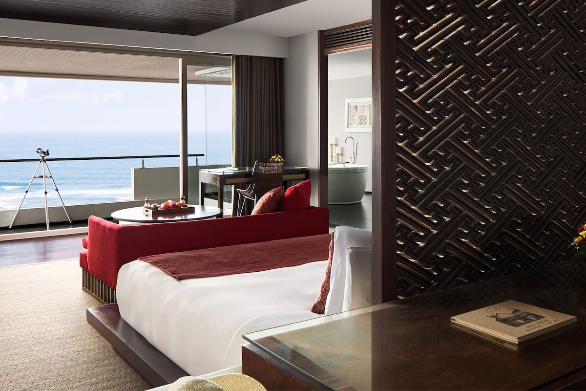 Quarto de hotel com cama, televisão, varanda vista para o oceano e secretária com luminária.