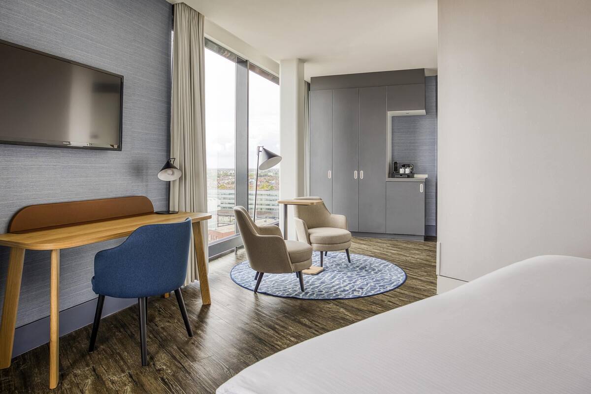 Quarto de hotel com cama, escrivaninha, duas cadeiras, uma janela ampla e uma cozinha compacta.