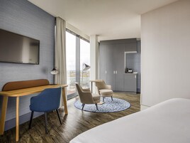 Quarto de hotel com cama, escrivaninha, duas cadeiras, uma janela ampla e uma cozinha compacta.