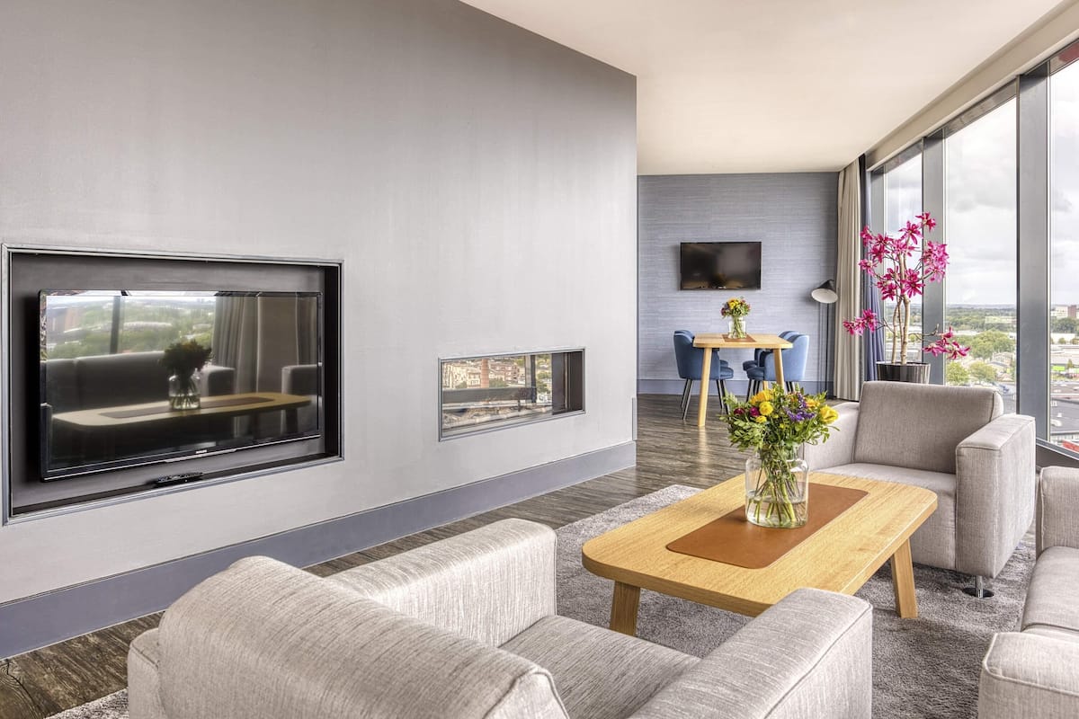 Uma sala de estar moderna com televisão de tela plana na parede, mesa de centro de madeira, sofás cinzas e vista da cidade através de grandes janelas.