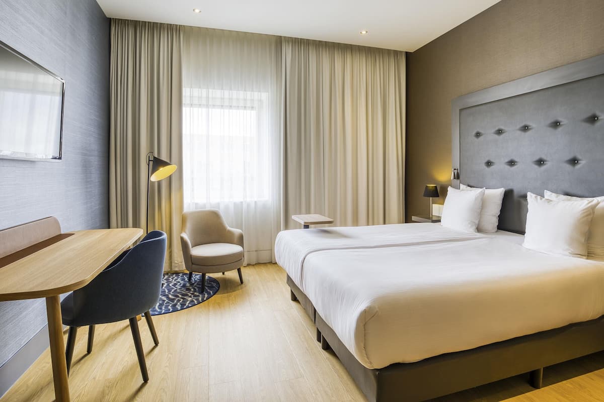 Quarto de hotel com uma cama grande, uma escrivaninha, uma cadeira, uma mesinha e um abajur.