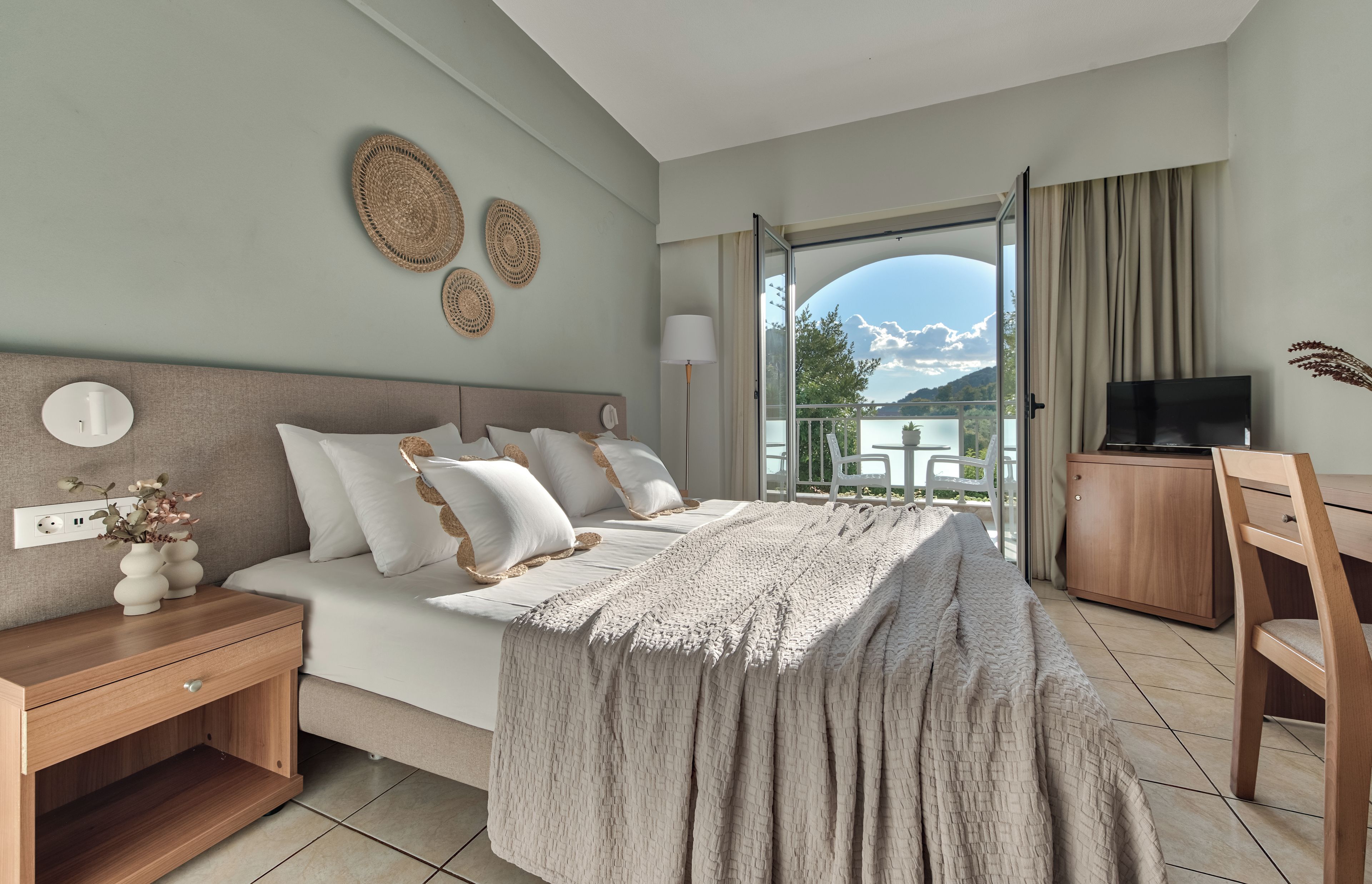 Quarto de hotel moderno com cama, mesa de cabeceira, luminária, TV e uma varanda com vista.