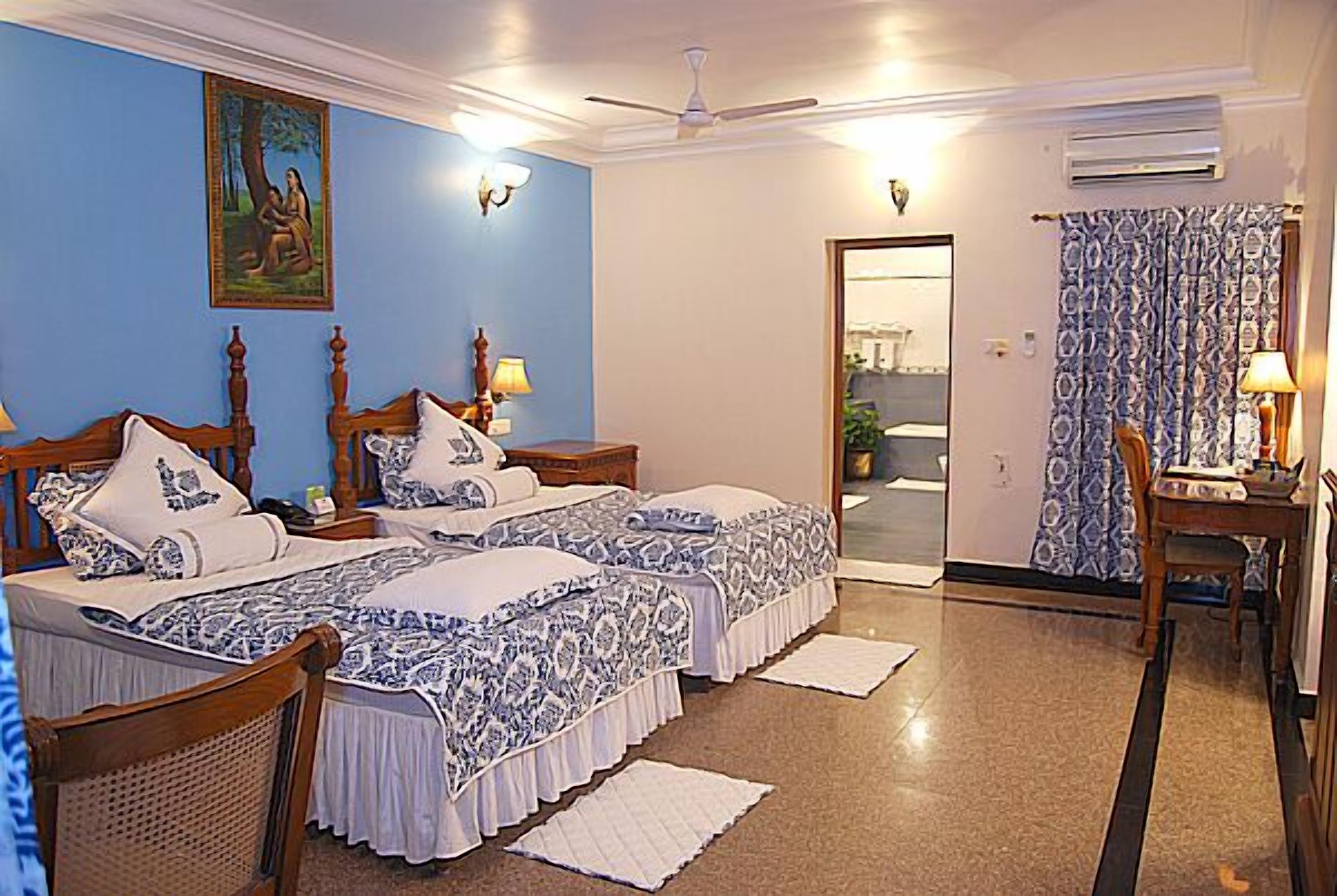 Quarto de hotel com duas camas, uma escrivaninha, uma cadeira, um ventilador de teto e uma porta que dá para uma varanda.